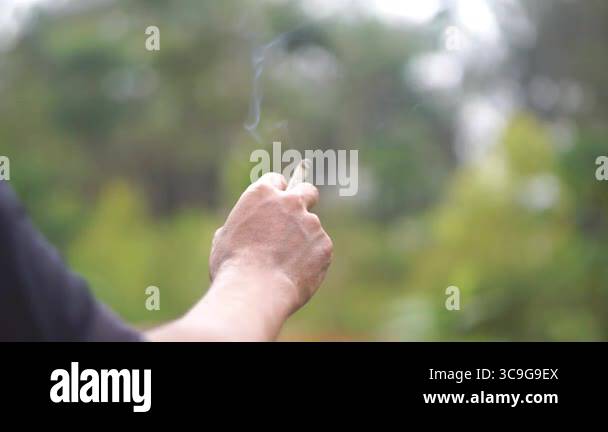 a man's hand holding a burning and smoking clove cigarette, outdoors in ...