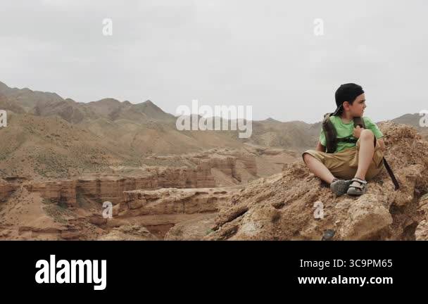 A small tourist boy in a cap and a backpack on his shoulders sits on ...
