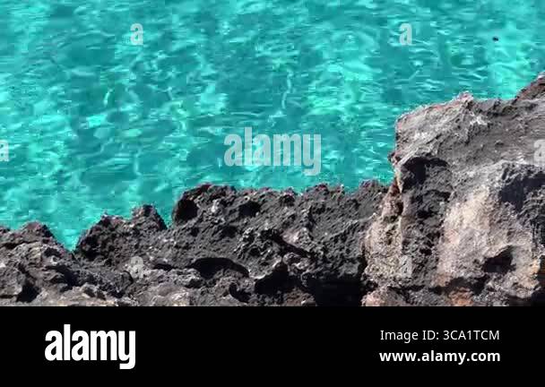 Clear turquoise water shimmering under sunlight, with rugged dark rocks ...