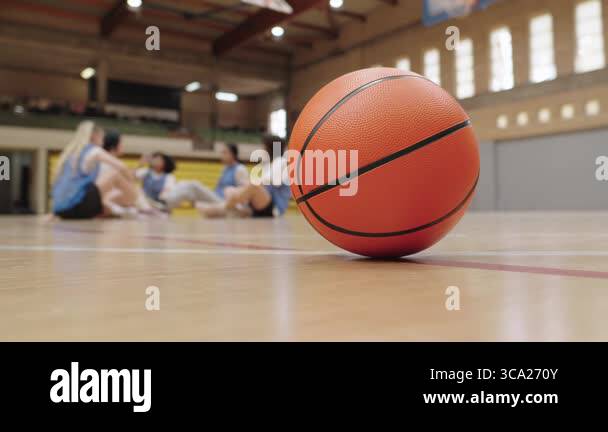 Basketball resting on hardwood court, female players recovering in ...