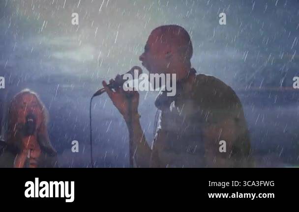 male singer extending arm toward audience on rain-soaked stage with ...
