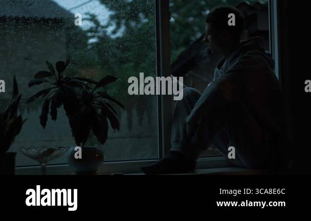 Young man sitting on window sill at rainy evening, smoking in silence ...