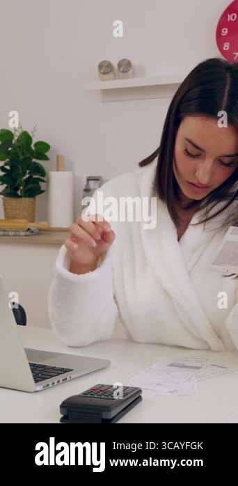 Vertical HD video of young pretty woman in bathrobe holding paper bill using calculator sitting ...