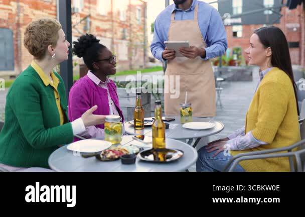 A diverse group of people enjoying a meal outdoors. A waiter takes an ...