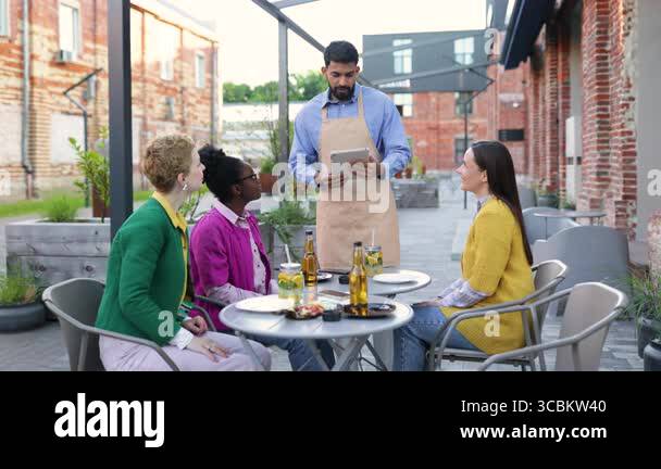 A diverse group of friends enjoys a meal at an outdoor cafe, served by ...