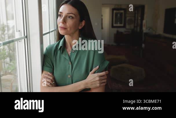 Serious sad young woman standing at window with arms folded at chest ...