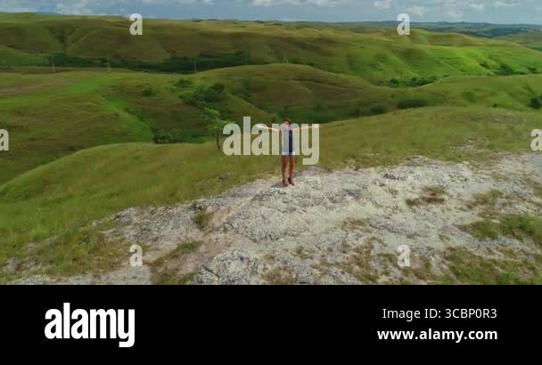 Young woman standing on top of a hill with open arms, embracing the beauty of rolling green ...