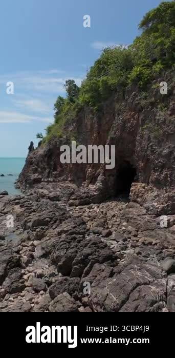 Vertical video. Drone Shot of Rugged Cliff Face with Shallow Cave Opening by Ocean and Thai Flag ...