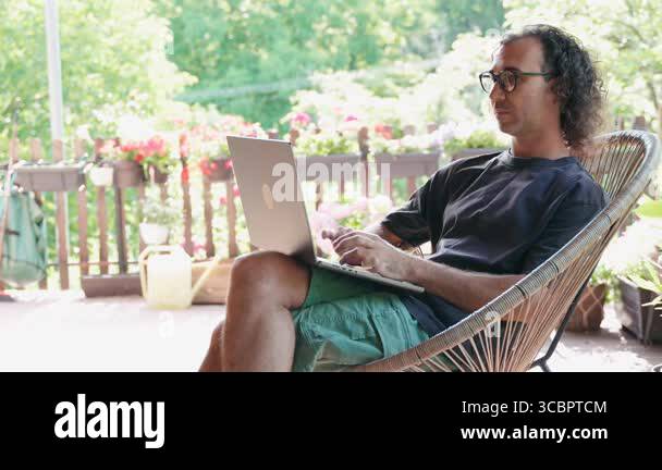 A young man is working on a laptop while sitting on the sunny terrace ...