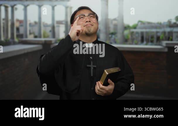 Asian priest with glasses and religious attire holds bible, gesturing ...
