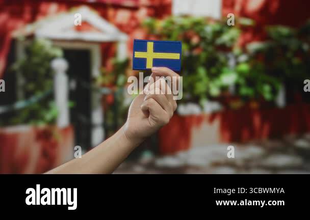Hand holding swedish flag outdoors in an urban city setting, focusing ...
