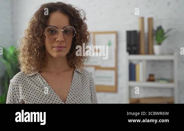 Woman standing confidently in modern office with curly hair and glasses ...