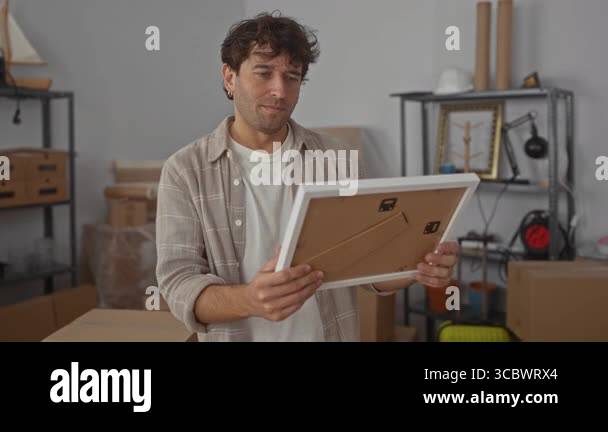 Handsome hispanic man holding picture frame in new home with boxes and ...