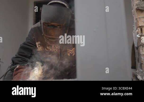 Welder in dark helmet bends toward glowing parts in silent workspace ...