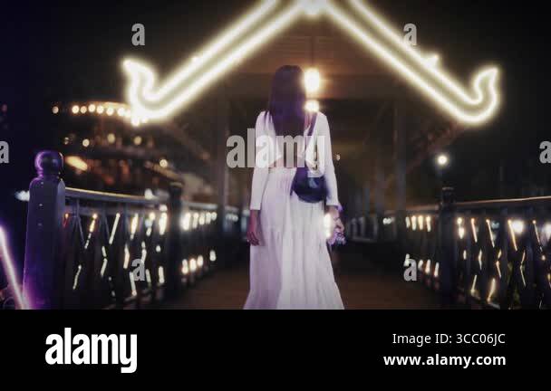 Back view of a woman walking alone on a lit bridge at night with a ...