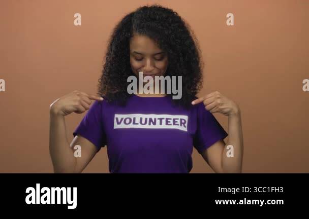 Young woman with curly hair in violet volunteer shirt smiles pointing at text against isolated ...