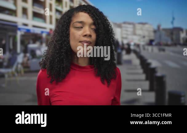 Woman wearing red shirt tilts head under bright sunlight on street with ...