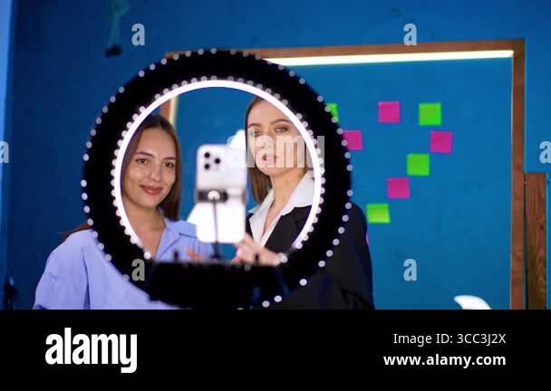 Two Caucasian ladies stand behind the camera with light ring. Recording ...