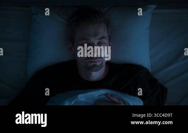 Close-up, in a dark bedroom at night, a man wakes up because of sleep ...