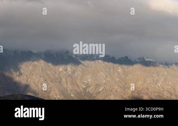 Clouds move over a rugged mountain range, casting dynamic shadows. The ...