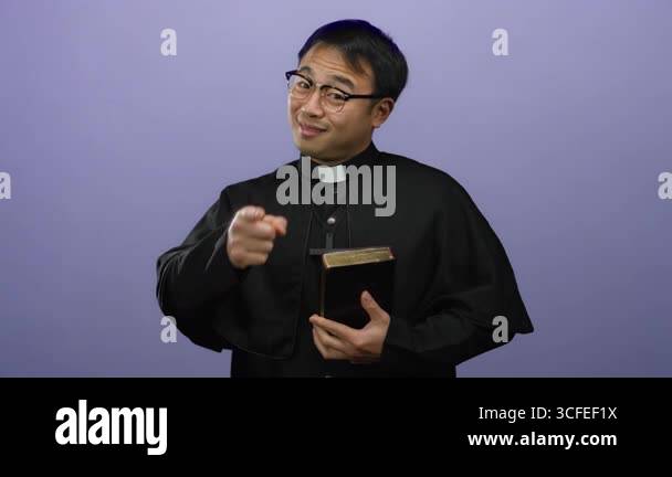 Asian man priest wearing glasses and black robe holds a bible against a ...