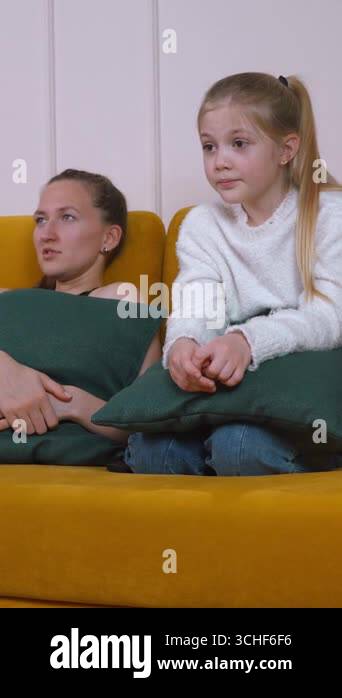 Mother and daughter sit together on a mustard yellow sofa, holding ...