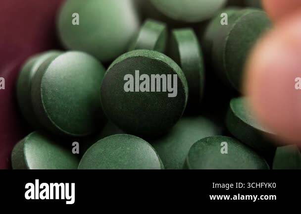 Green spirulina algae tablets in red ceramic bowl and fingers taking ...