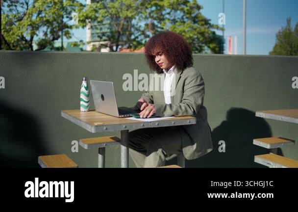 A trans woman in professional attire works outdoors using a laptop ...