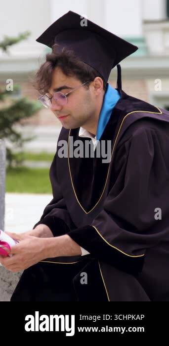 Young man sits on bench gazing thoughtfully at diploma. Student looks ...