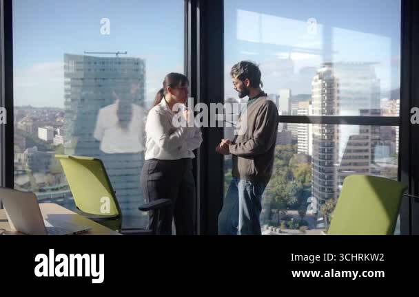Two colleagues standing by a large office window having a casual ...