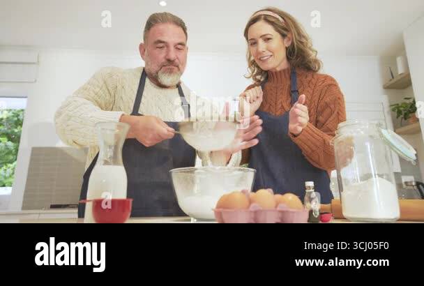 Couple tapping sieve and sifting flour into mixing bowl then pouring milk, cracking eggs for ...