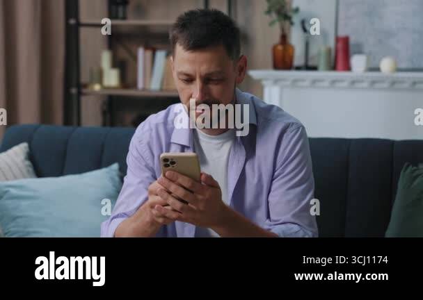 Attractive smiling happy young man use phone sitting on sofa at home ...