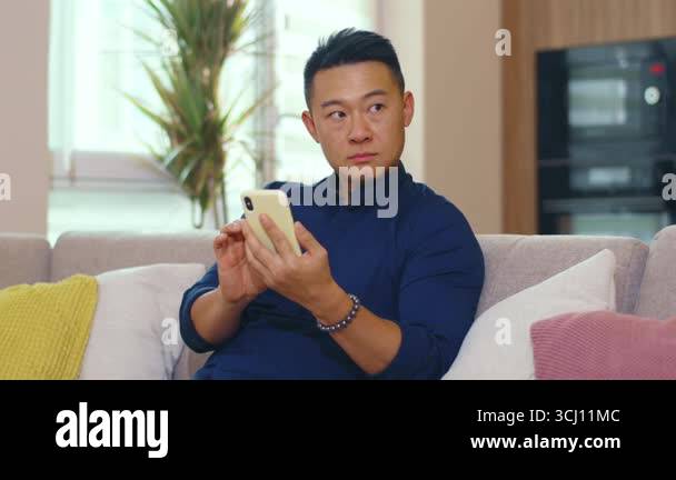 Serious young asian man use phone sitting on sofa look around at home ...