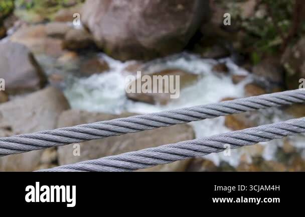 A sturdy steel cable stretches across a rocky river in a forest setting ...
