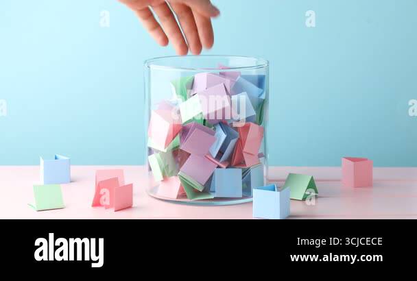 Woman picking color paper piece from glass jar at pink wooden table ...