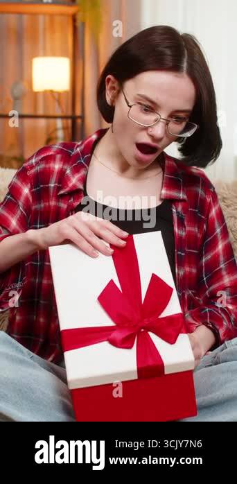 Young happy woman opens present on home sofa, smiling with surprise and ...