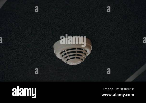 A white smoke detector is attached to a textured black ceiling. The ...