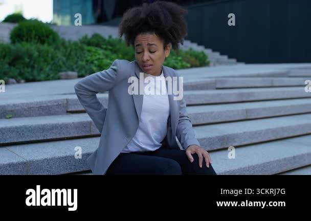 Young woman in gray blazer sits on stone steps holding her lower back ...