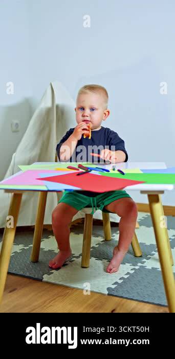 Cute toddler in a T-shirt sits at home at a children's table and draws ...