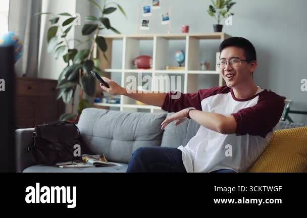 attractive young chinese man with glasses sits on the couch dancing and ...