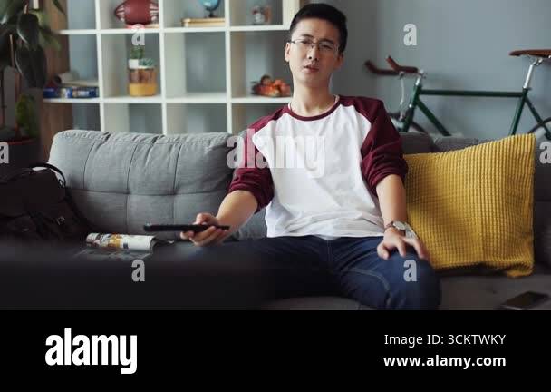 Young attractive chinese man with glasses sits on the couch and turns ...