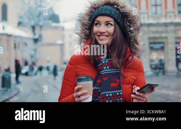 Beautiful happy woman walks around the city drinking coffee using the phone in winter smiling ...