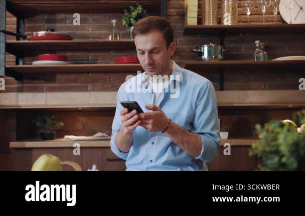 Handsome sociable young man using a smartphone sitting in the kitchen ...