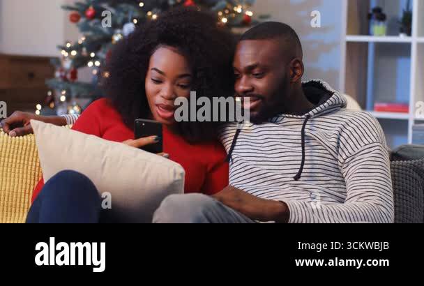 Nice African couple sitting on comfortable sofa in hall of big house. Young people looking on ...