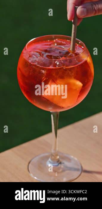 hand mixing a glass of classic red spritz cocktail with ice and straw ...
