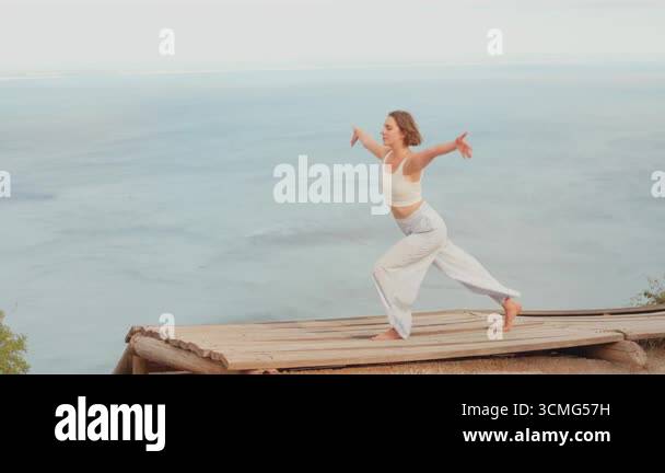 Young woman practicing yoga asanas on wooden platform by ocean. She ...