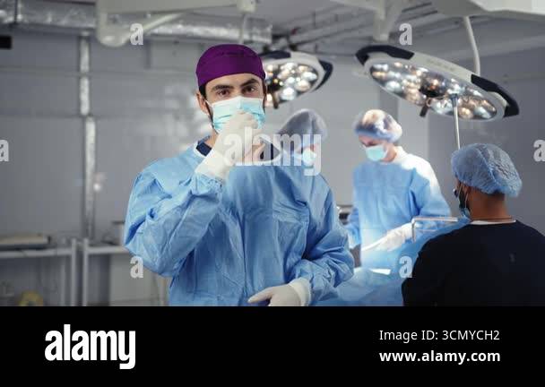 A surgeon in a blue surgical gown and cap smiles in an operating room ...
