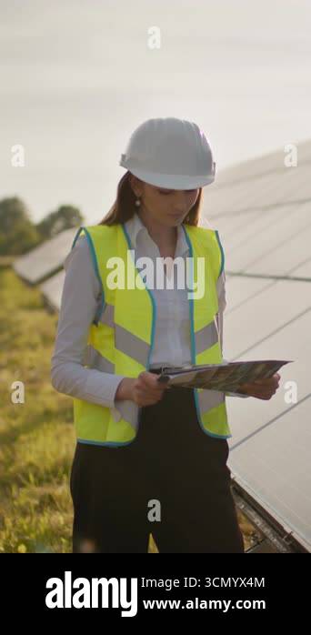 A female engineer in a safety vest inspects solar panels at a solar farm, ensuring efficient ...
