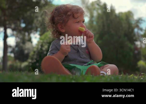 Curly-haired toddler eating snack while sitting on grass in park with ...