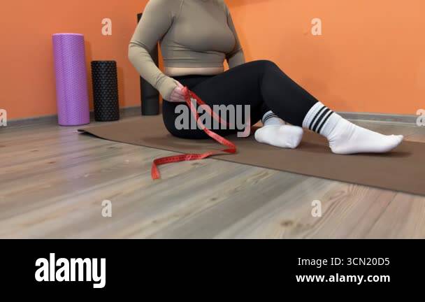 A woman sits on a mat at home measuring her thigh with a tape. She ...
